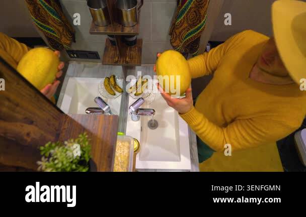 A young man washes a melon fruit under a water tap in an apartment. A ...