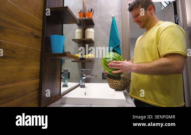 A young man with a smile washes a watermelon under the water tap in the ...