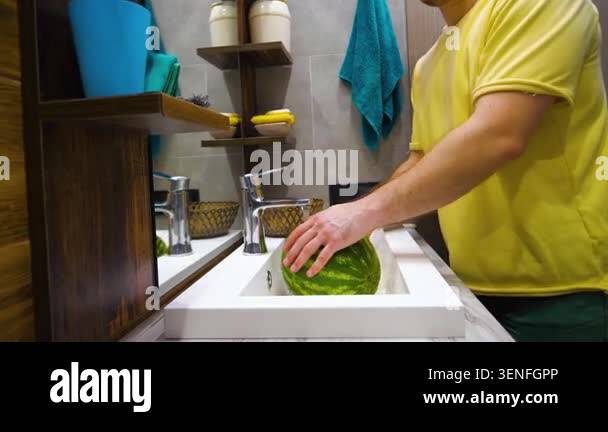 A young man washes a watermelon in the kitchen, washing the peel of a ...