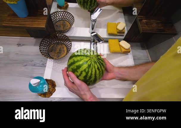 A man washes a watermelon under a tap with water, top view. Wash a ripe ...