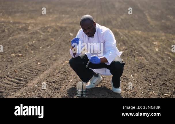 Male scientist in a white lab coat and gloves crouching in a plowed ...