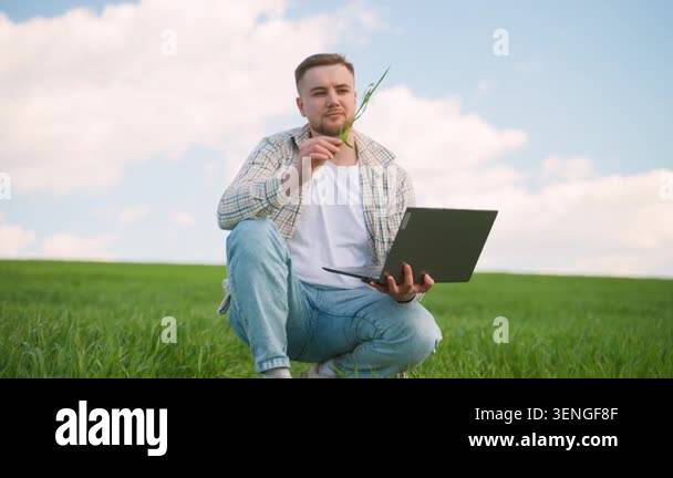 Young agronomist squatting in a green field, holding a laptop and ...