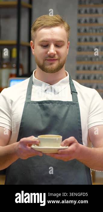 Cheerful male barista in an apron holding a freshly made cup of coffee ...