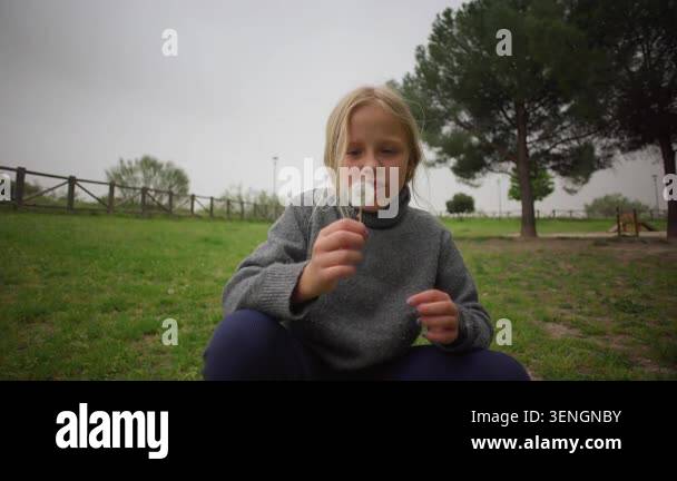 Blonde girl in grey sweater sitting on grass and blowing fluffy ...