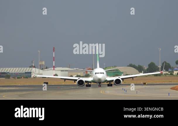 Front view of a narrow body jet taxiing along the runway with hangars ...