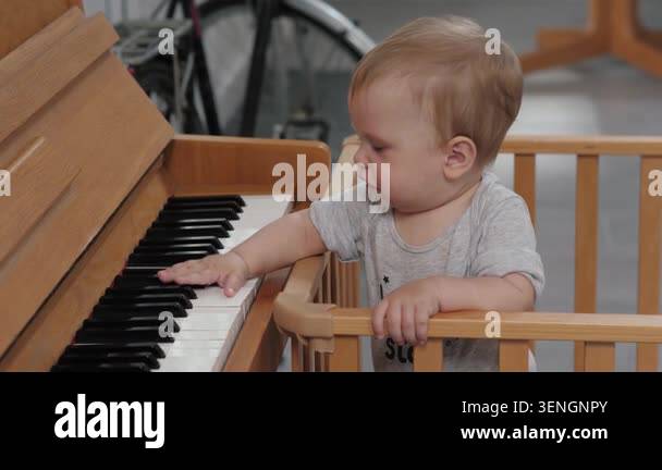 Baby explores piano keys. Young child stands in wooden playpen examines ...