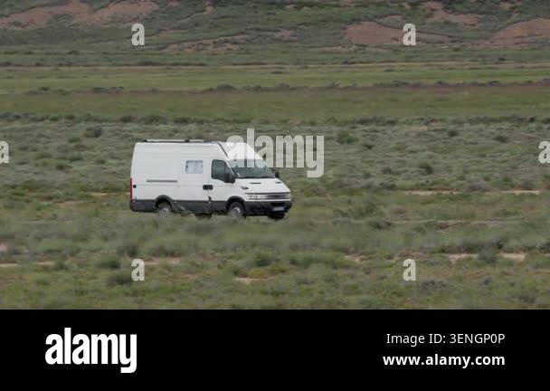 White camper van drives on rural road crossing green grassy field with ...
