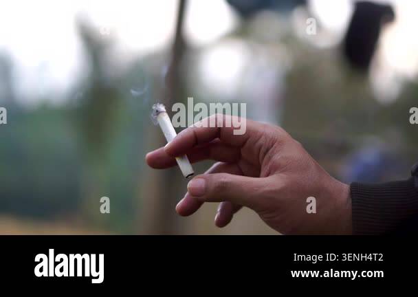 Close-up shot of a person's hand holding a lit white cigarette outdoors ...