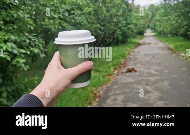 Hand with a paper cup of coffee on the background of a green park alley ...