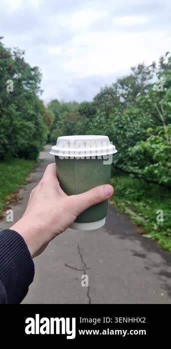 Hand with a paper cup of coffee on the background of a green park alley ...