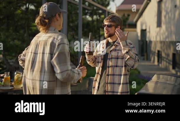 Two adult men stand outside in warm evening light, talking casually ...