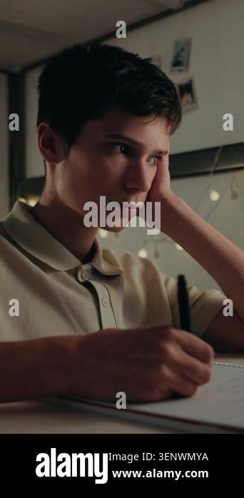 A pensive teenage boy sits at his desk at night, writing in a notebook ...