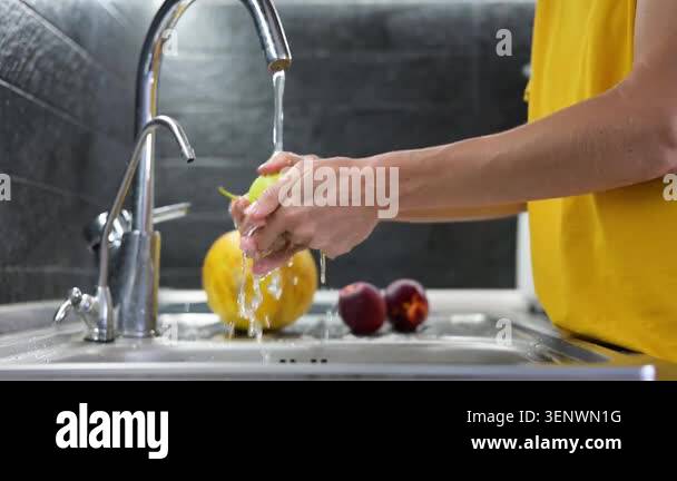 Close up of hands rinsing fresh ripe pear beneath flowing tap water in ...