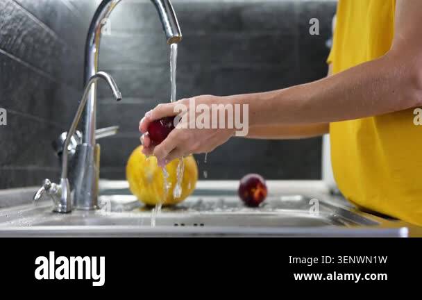 Close up of hands cleaning fresh red plum beneath flowing tap water in ...