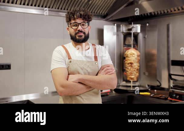 Confident male chef wearing an apron and glasses, standing with arms ...