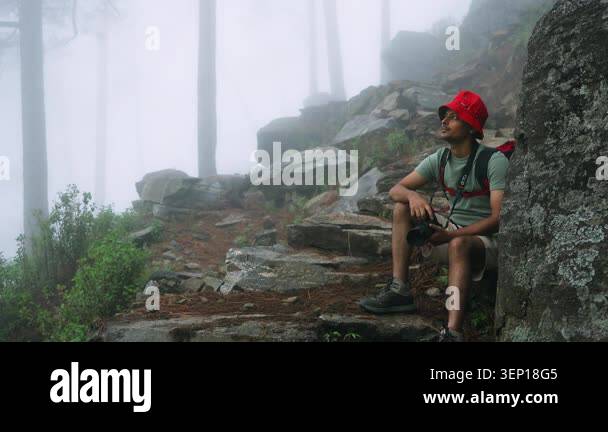 A lone trekker sits on a rugged mountain path surrounded by dense ...