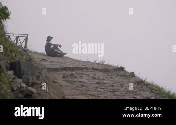 A lone trekker sits on a rugged hillside trail surrounded by dense ...