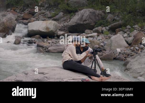 A trekker sits beside a flowing river with a camera tripod in the ...