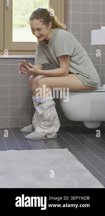 Vertical video of a smiling young woman sitting on the toilet and ...
