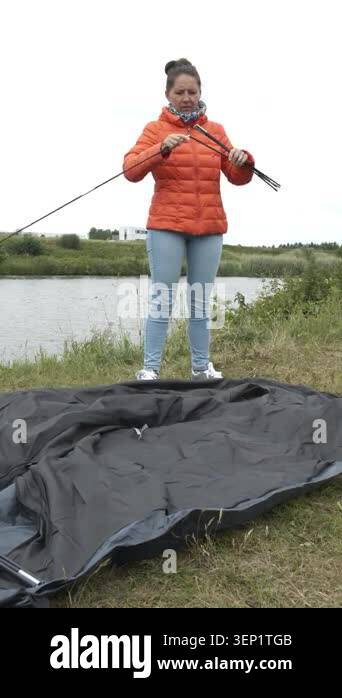 A woman in an orange jacket connects tent poles by a calm lake. She ...
