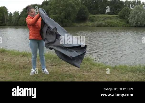 A woman unfolds a dark gray pop up fishing shelter on a grassy bank ...