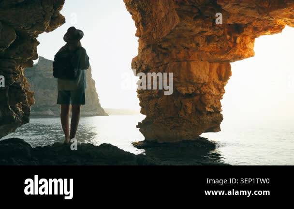 young man in white dress and hat standing by sea on rocky cliff and ...