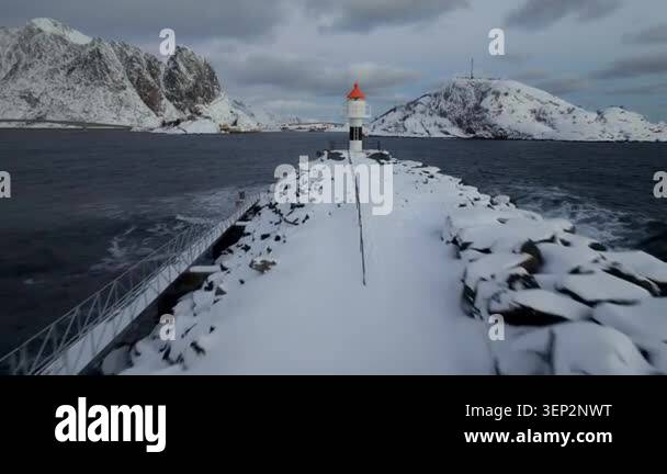 A snow-covered pier extends into a dark, icy sea with a small ...