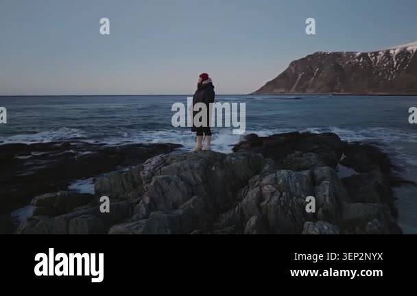 A lone figure in a dark parka, white boots, and a red hat stands on ...