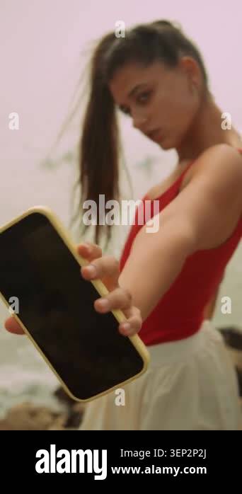 Young woman arching back with smartphone extended by rocky ocean ...