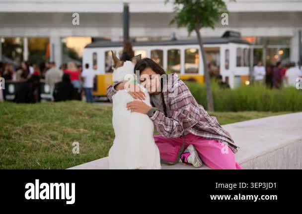 A young woman sits outdoors and gently hugs a white dog near a blurred ...