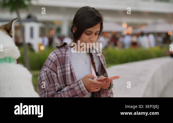 A young woman stands outdoors in a park at dusk, looking down at her ...