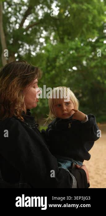 Mother holding toddler in woods, observing quietly, green canopy ...