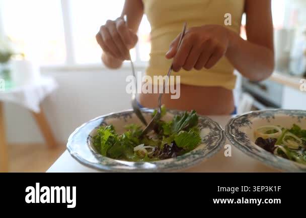 An adult woman serves fresh salad onto plates in a bright home kitchen ...