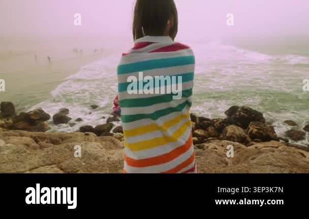 White woman facing ocean on rocky jetty, wrapped in colorful striped ...
