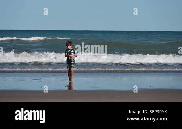 Young boy standing at the water edge on a beach, holding a bucket, with ...