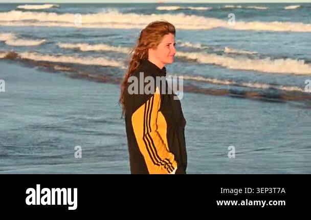 A woman with long, wavy hair walking along a beach with waves crashing ...