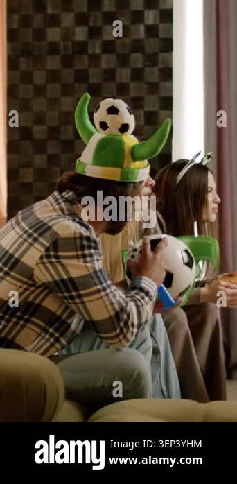 Friends in festive hats enjoy a sports event, holding inflatable props ...
