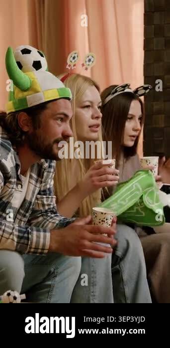 Friends in festive hats enjoy a sports event, cheering and holding ...
