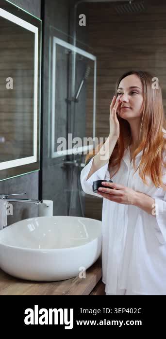 Young woman in white robe applying moisturizing cream on face in ...