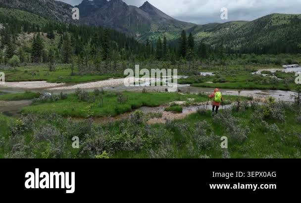 A lone hiker with a bright backpack walks along a shallow river in a ...