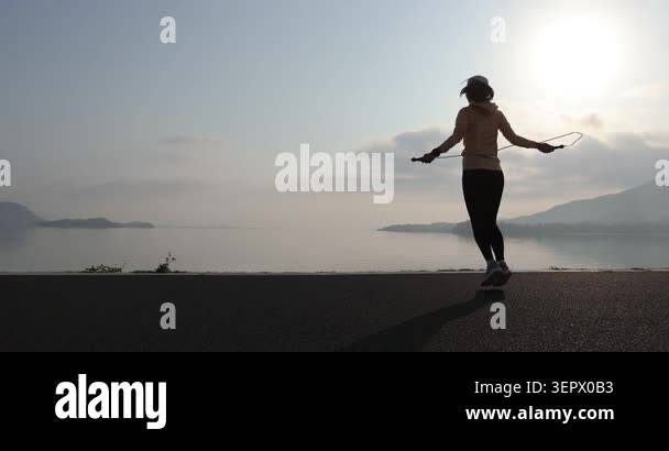 Silhouette of a woman jumping rope by a lake at sunrise, with mountains ...