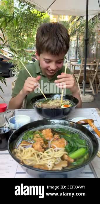 A child is smiling while holding chopsticks and eating noodles at a ...