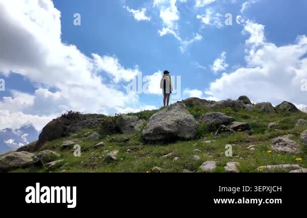 A person with a backpack stands on a rocky hill under a cloudy blue sky ...