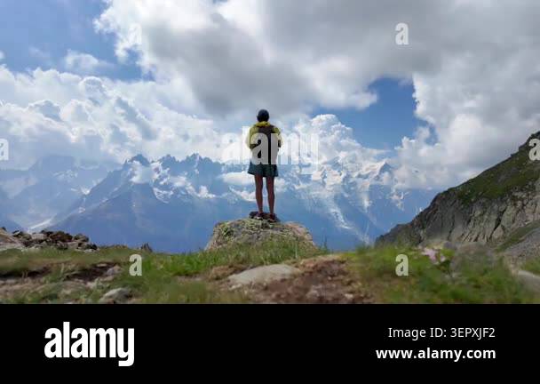 A lone hiker stands on a rock, looking out at a vast mountain range ...