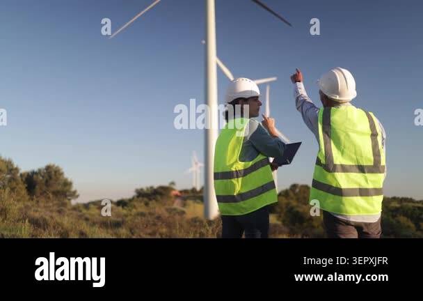 Two engineers in hard hats and safety vests stand in a field of wind ...