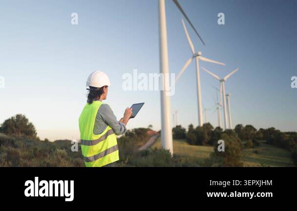 A woman in a hard hat and safety vest uses a tablet near wind turbines ...