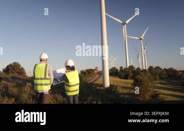 Two engineers wearing hard hats and safety vests review blueprints at a ...