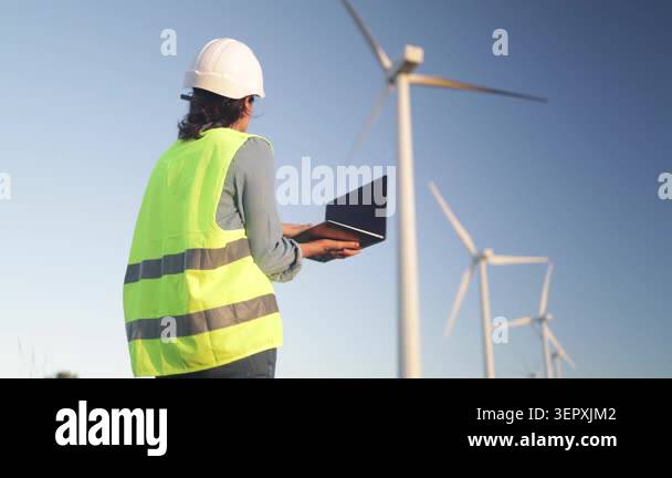 A woman wearing a hard hat and safety vest holds a laptop while ...