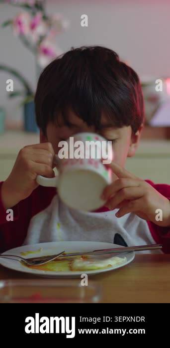 A young boy drinks from a cup while eating breakfast at the table ...