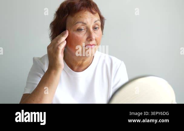 Adult woman examines wrinkles in a small mirror indoors. An older woman ...